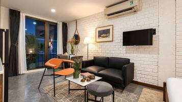 Uma sala de estar moderna com um sofá de couro preto, uma mesa de centro redonda e uma poltrona laranja. Há uma TV de tela plana na parede e um vaso de planta na mesa.