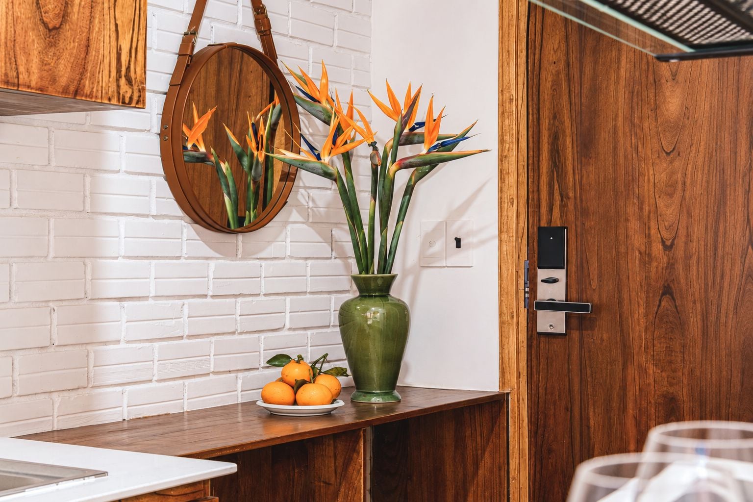 Uma bancada de cozinha com um vaso verde contendo flores laranjas, um prato com laranjas e um armário de madeira.