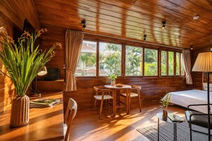 Interior de uma cabana de madeira com área de jantar, cama e amplas janelas com vista para uma paisagem tropical.
