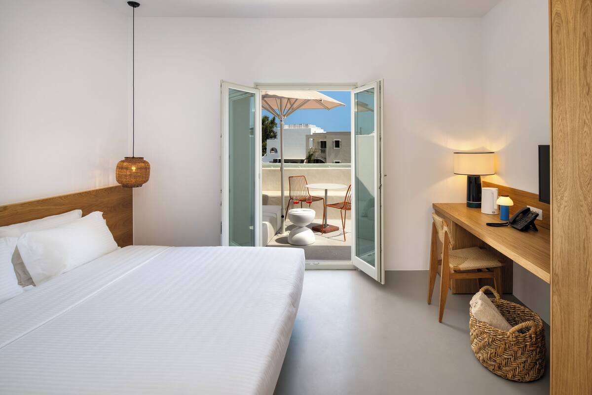 Quarto de hotel com cama, escrivaninha com luminária, cesta de vime e vista para área externa com mesa e cadeiras.