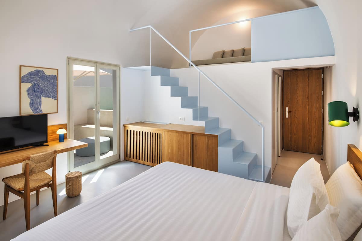 Quarto de hotel moderno com cama, escrivaninha com TV, escada e porta.