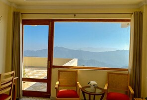 Quarto com uma grande janela com vista para a montanha, duas cadeiras de vime com almofadas vermelhas e uma pequena mesa com um vaso.