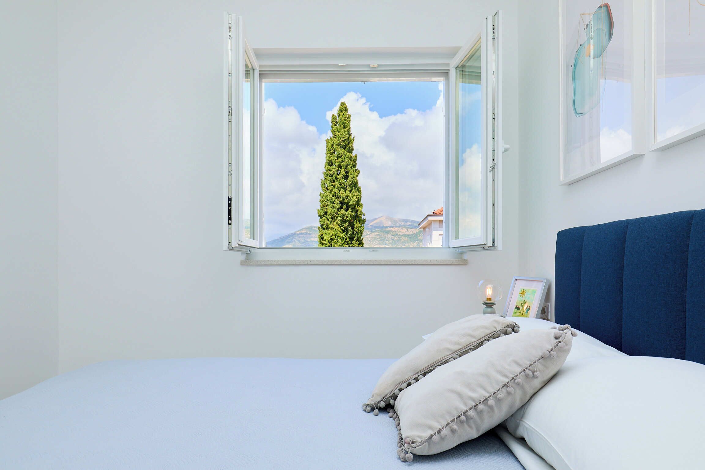 Quarto com cama, almofadas, janela com vista para uma árvore e montanhas, e luminária fixada na parede.
