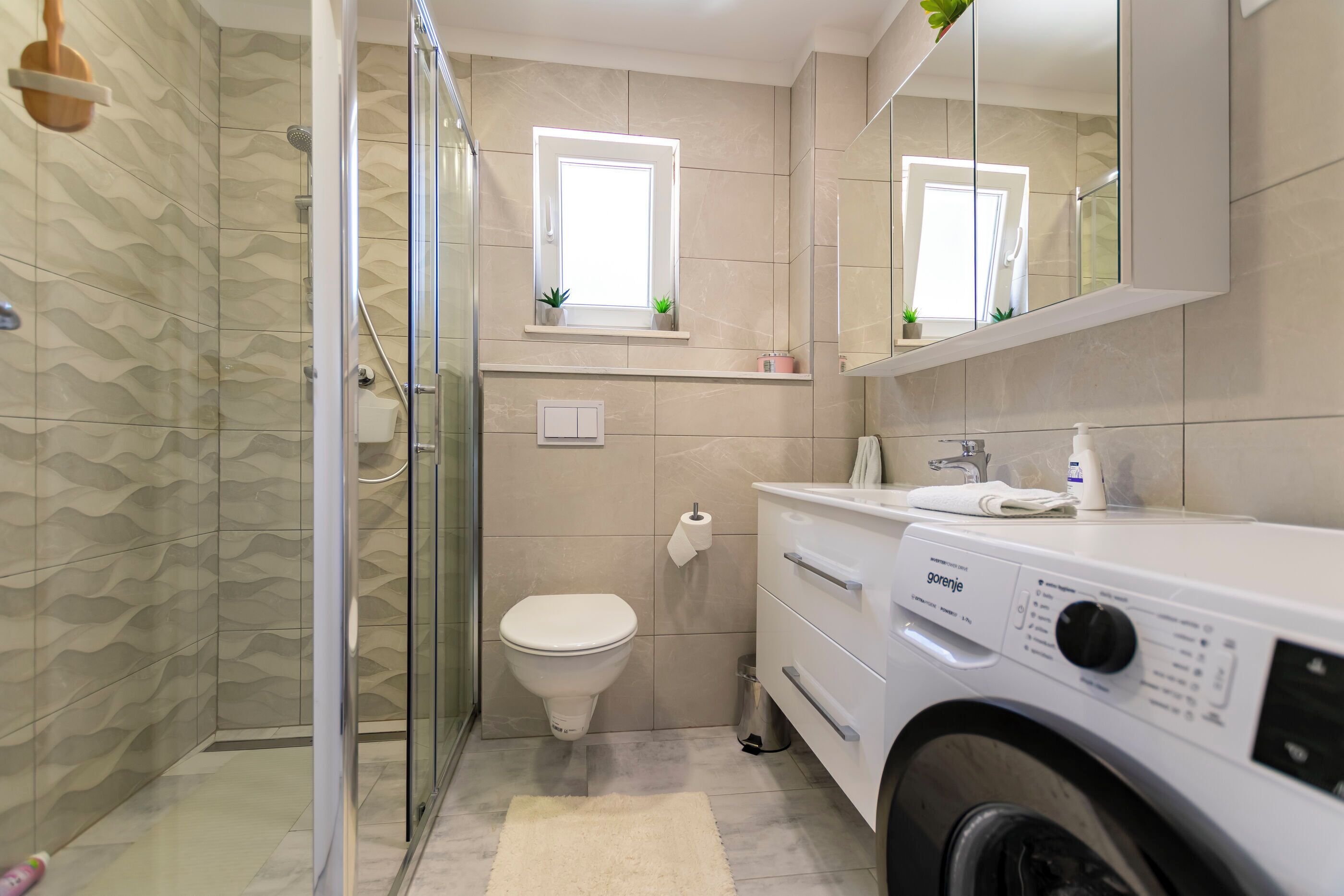 Uma casa de banho moderna com cabine de duche em vidro, sanita, máquina de lavar roupa e espelho sobre a lavatório.