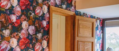 Um quarto com papel de parede floral, uma porta de madeira, uma cama com almofadas, um abajur e uma janela com vista.