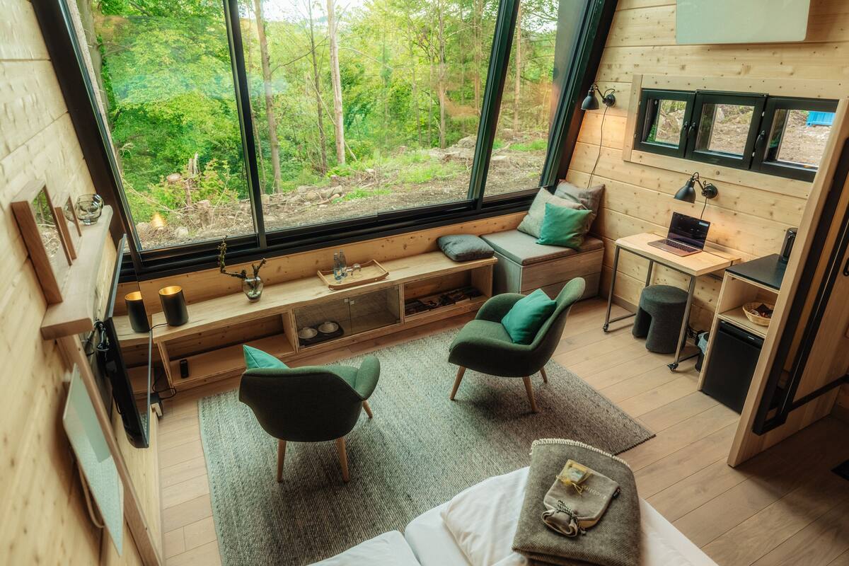 Interior acolhedor de uma cabana com uma janela grande que oferece vista para a floresta, uma área de estar com um laptop e uma pequena cozinha compacta.