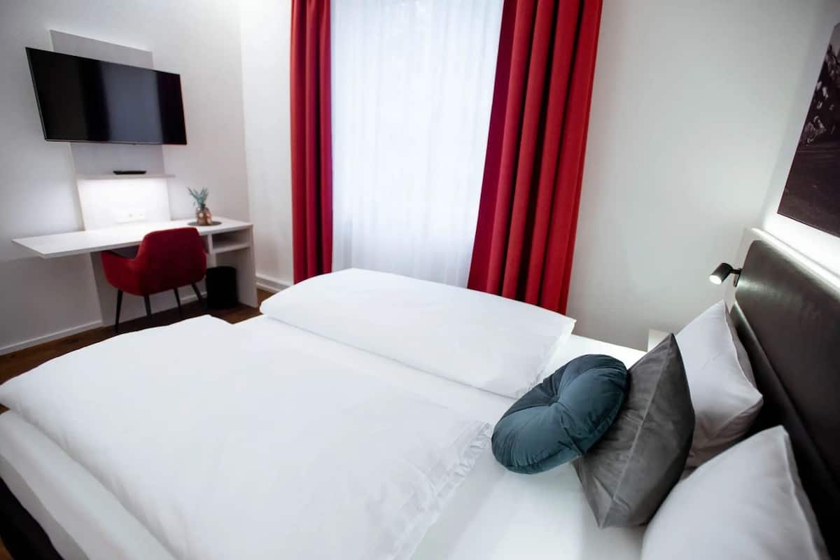 Quarto de hotel com uma cama, uma mesa, uma cadeira, uma televisão e cortinas vermelhas.