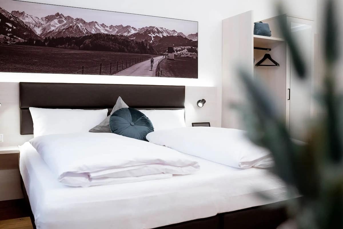 Cama bem arrumada com lençóis brancos e cabeceira escura. Uma pintura de paisagem montanhosa é visível acima da cama.