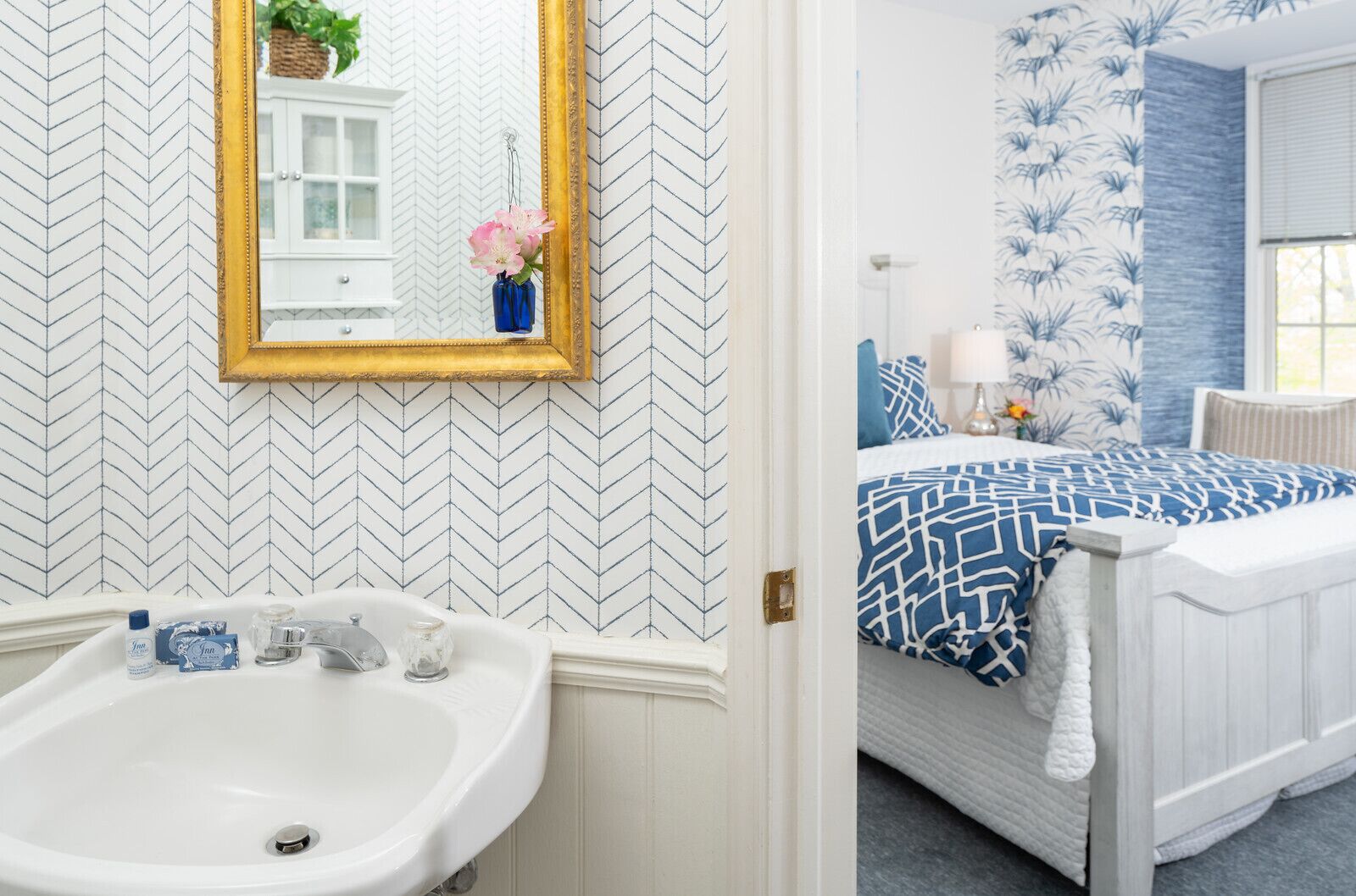 Casa de banho com uma pia branca, um espelho com moldura dourada e papel de parede estampado.