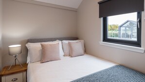 Quarto com cama, mesa de cabeceira, luminária e janela com vista para uma casa.