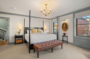 Uma cama com dossel, um banco, um candelabro, um retrato emoldurado e vista da rua pela janela.