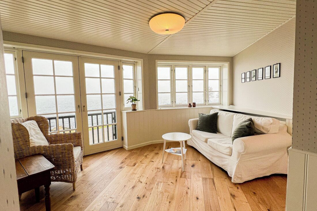 Quarto com sofá branco, cadeira de vime, mesa pequena e janelas amplas com vista para a água.