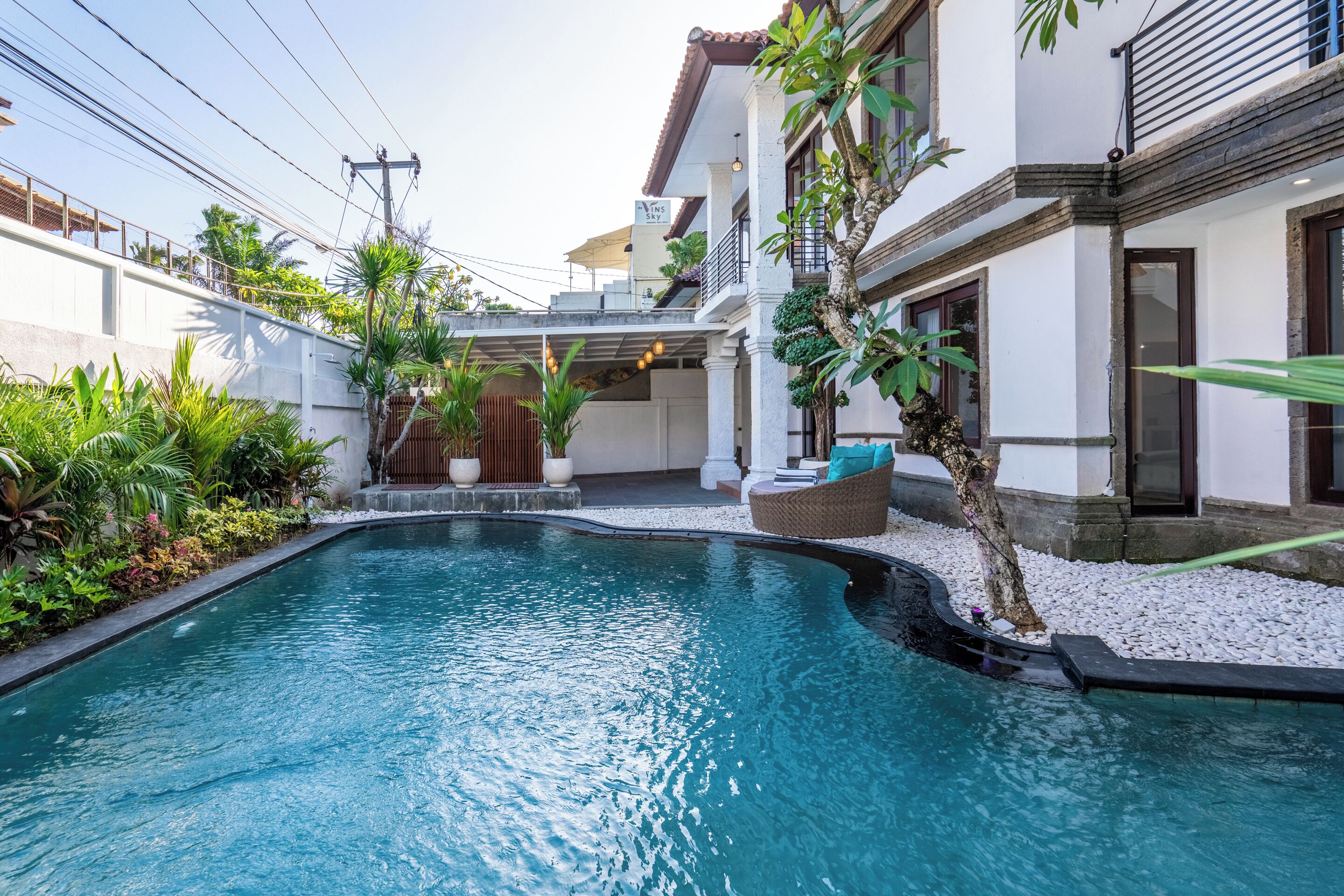 Piscina com parede branca e varanda, cercada por plantas e um edifício com a placa 'FUS SKY'.