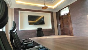 Uma sala de conferências com uma mesa comprida, cadeiras pretas e uma televisão fixada na parede.