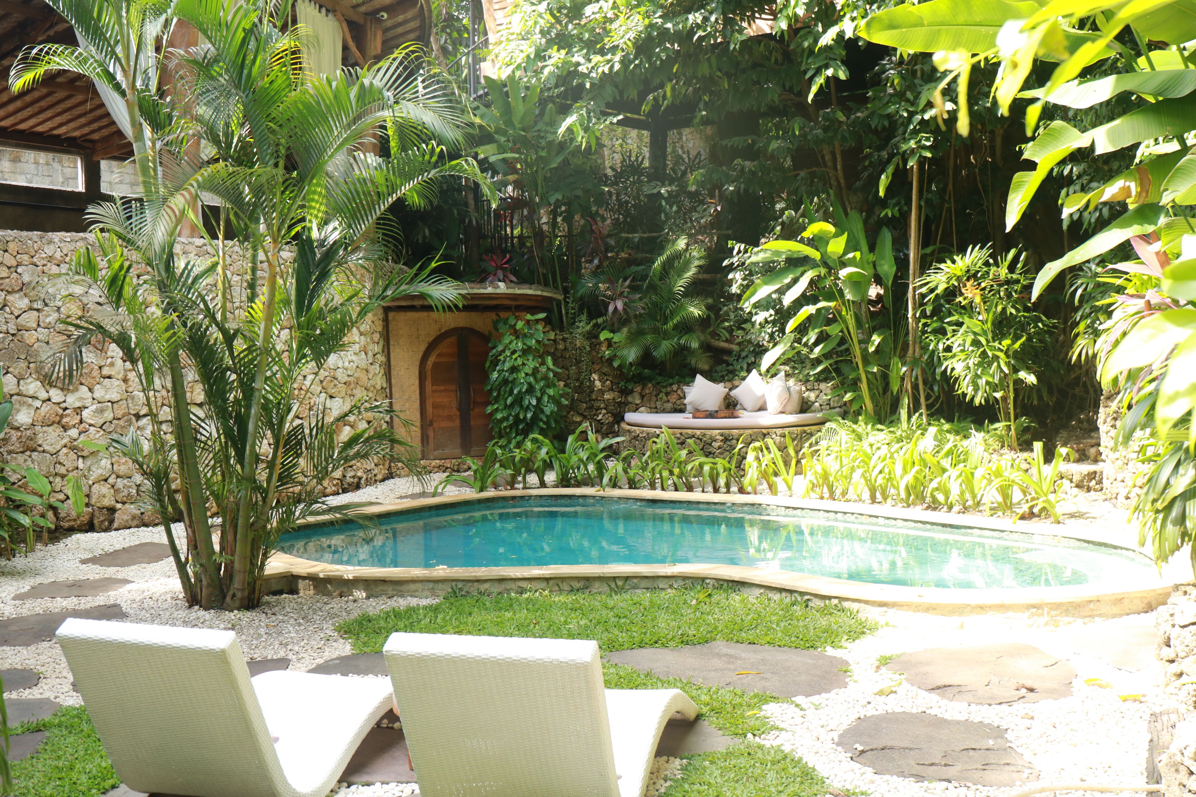 Piscina rodeada por vegetação, paredes de pedra e duas cadeiras de descanso.