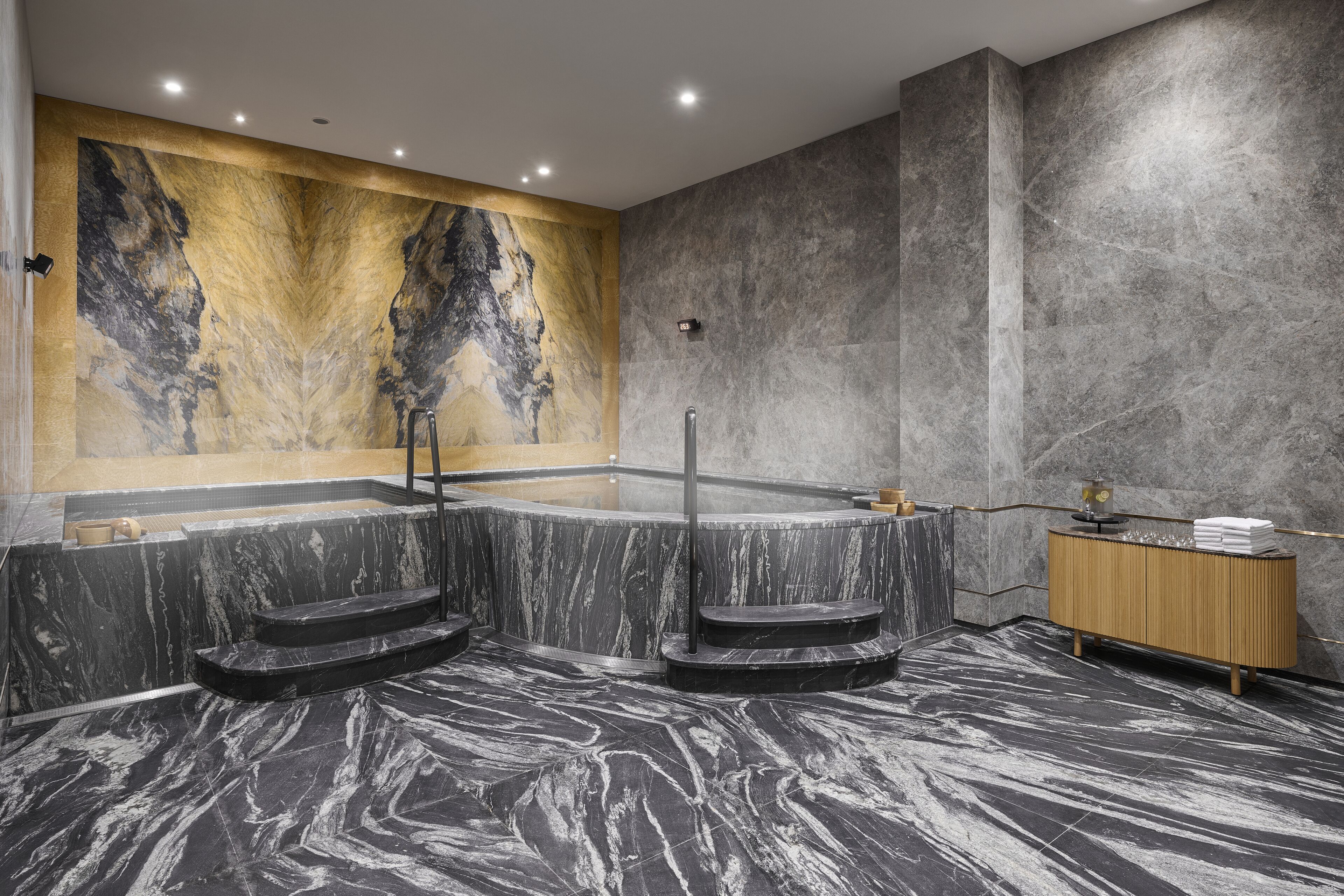 Uma área de spa moderna com piso de mármore, uma banheira grande fixada na parede e um espelho com bancada de madeira e toalhas.