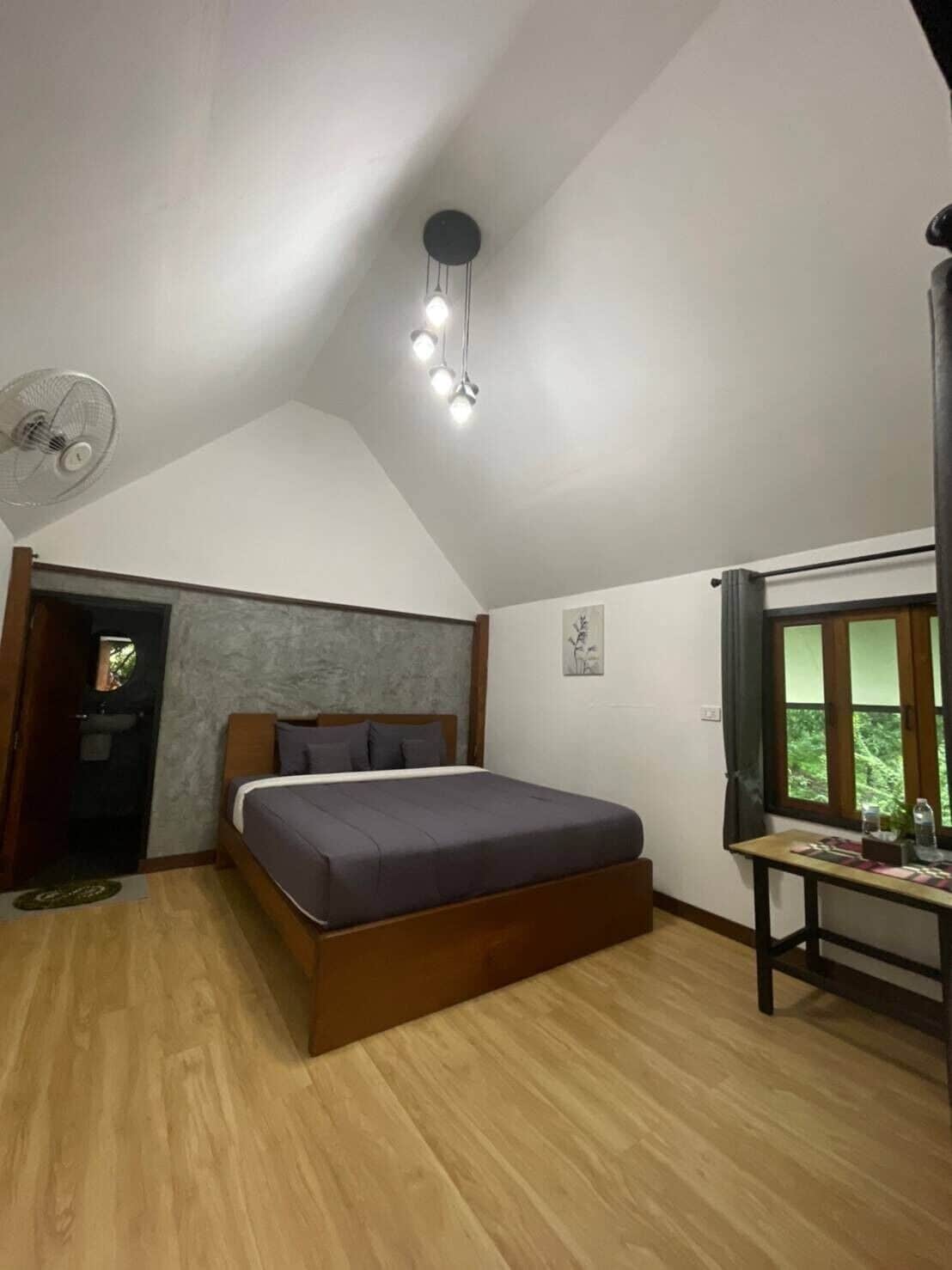 Quarto com teto inclinado, cama com estrutura de madeira, ventoinha no teto e janela com vista para vegetação.