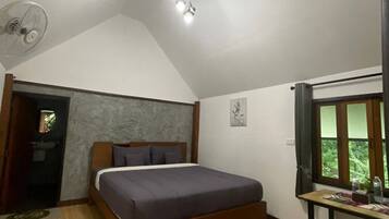 Quarto com teto inclinado, cama com estrutura de madeira, ventoinha no teto e janela com vista para vegetação.