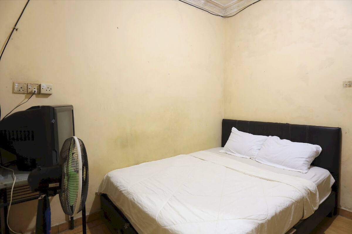 Quarto pequeno com uma cama de solteiro, parede simples, uma escrivaninha com computador e um ventilador.