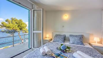Quarto com uma janela grande, uma cama com colcha estampada, duas almofadas, uma mesa de cabeceira com abajur e vista para o mar.