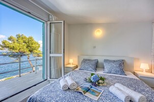 Quarto com uma janela grande, uma cama com colcha estampada, duas almofadas, uma mesa de cabeceira com abajur e vista para o mar.