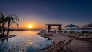 Uma área de piscina tranquila com espreguiçadeiras e guarda-sóis, com vista para um pôr do sol sobre a água.