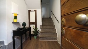 Uma escadaria que leva a um segundo andar, uma porta de madeira, um aparador preto com uma planta em vaso e uma prateleira na parede com um vaso amarelo.
