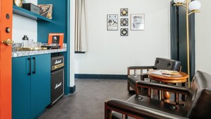 Espaço compacto com armários cor turquesa, máquina de café, amplificador de guitarra, mesa de jantar com cadeiras, e parede decorada com quadros e um toca-discos.