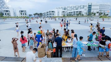Uma praça ampla com edifícios modernos ao fundo, um grupo de pessoas reunido em volta de uma mesa com diversos objetos, e algumas pessoas sentadas em bancos.
