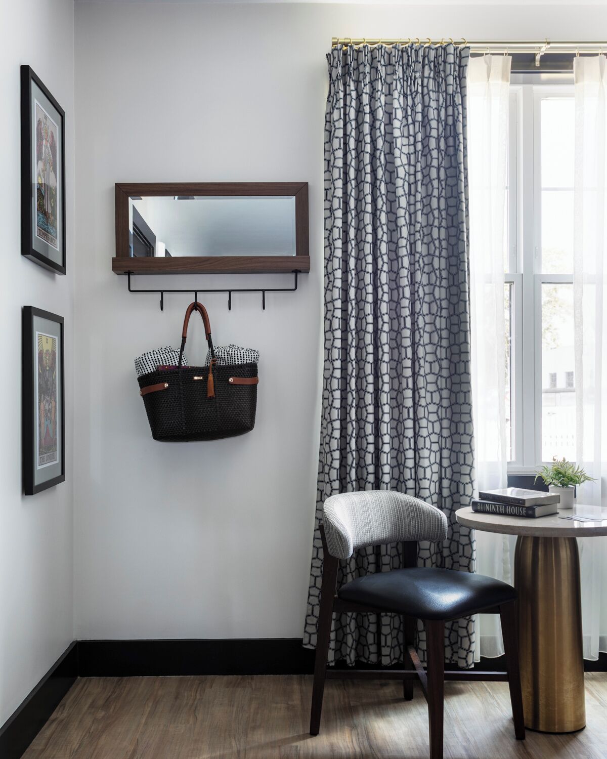 Quarto moderno com piso de madeira, cortina estampada em preto e branco, cadeira de madeira, mesa redonda pequena com uma planta e espelho fixado na parede.