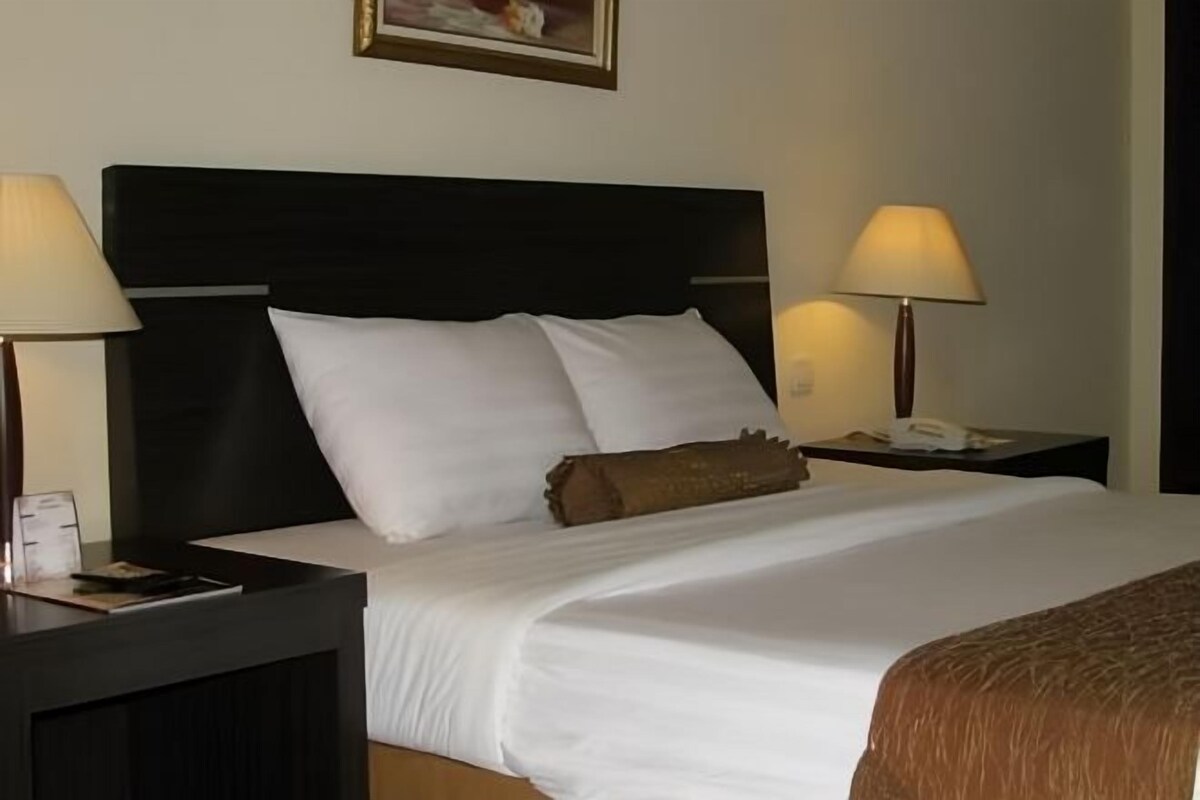 Cama bem arrumada com lençóis brancos, cabeceira escura e duas luminárias laterais.