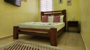 Quarto com cama de madeira, mesa de cabeceira com luminária, TV fixada na parede e janela com persianas.