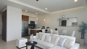 Uma sala de estar moderna com um sofá branco, uma mesinha de apoio preta e uma cozinha ao fundo.