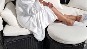Uma pessoa sentada em uma cadeira de descanso externa, com capa branca e almofada preta, segurando uma xícara preta.