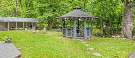 Um gazebo em um ambiente externo exuberante e verde, com uma área de varanda coberta ao fundo.