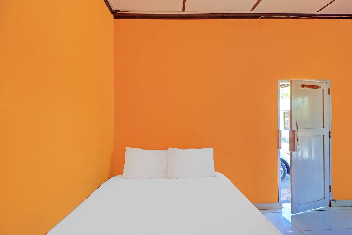 Quarto com parede laranja, cama branca com duas almofadas e porta que dá para o exterior.