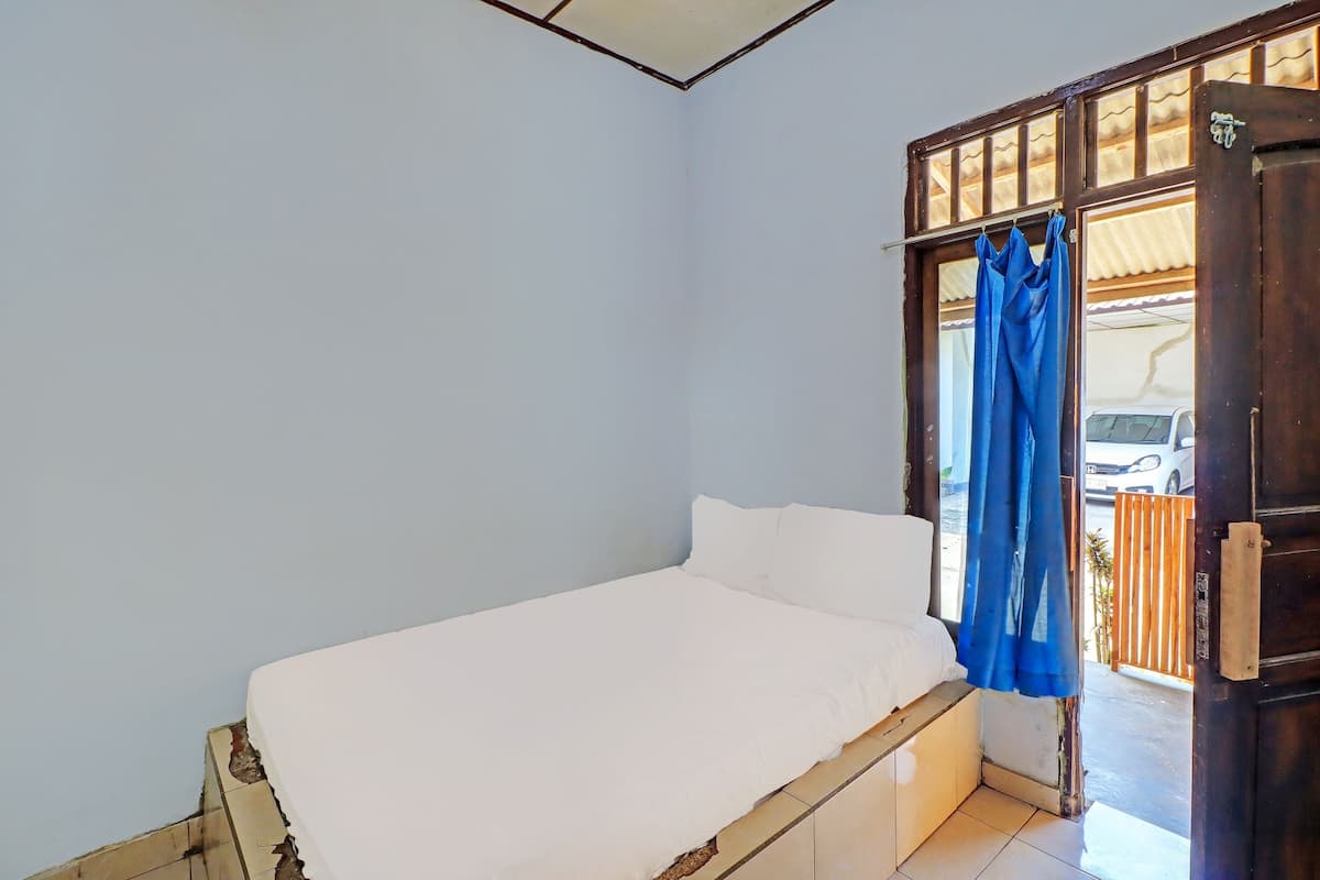 Quarto pequeno e simples com uma cama, uma janela com cortinas azuis e vista para um carro estacionado lá fora.