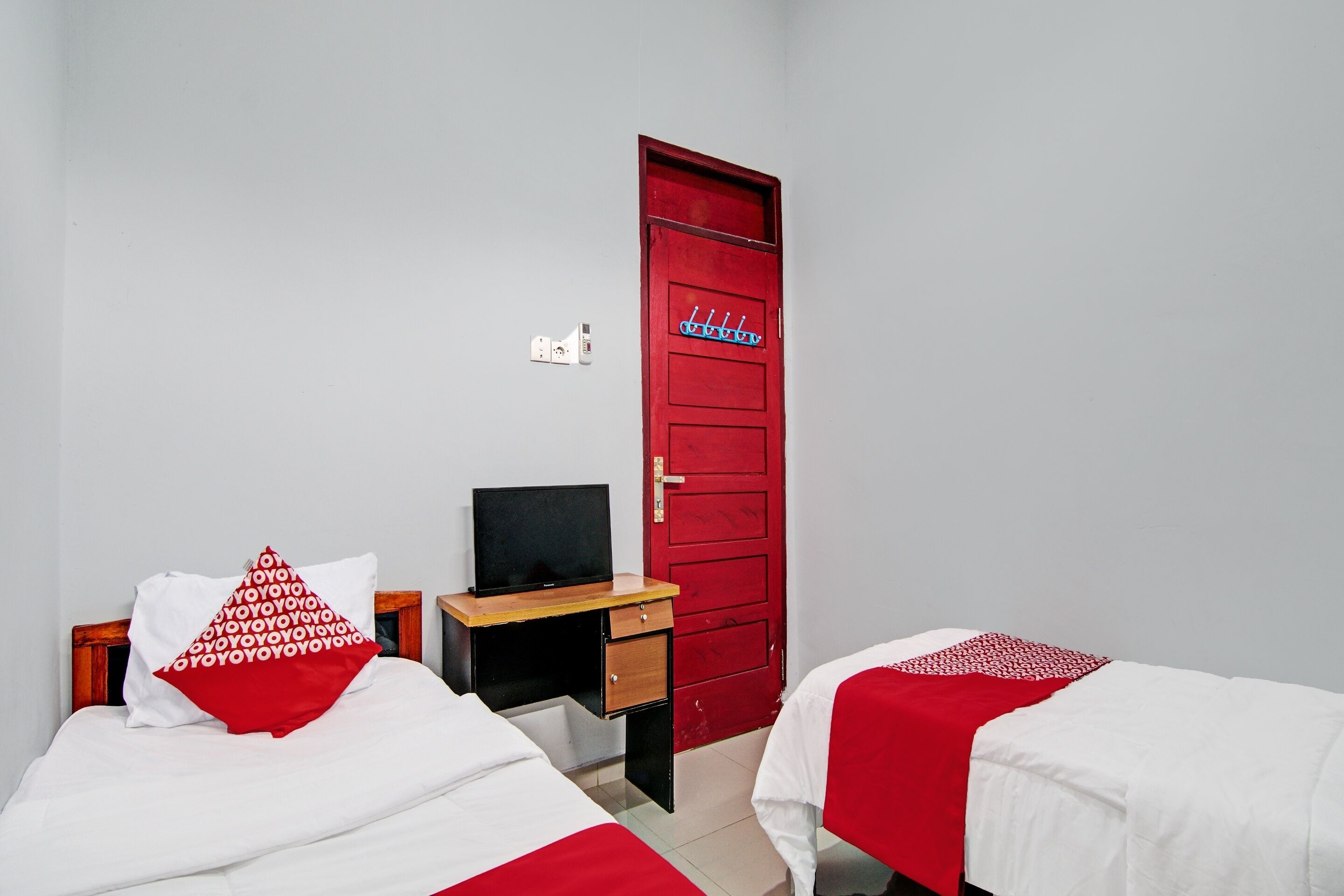 Quarto com duas camas, uma escrivaninha com televisão, uma porta vermelha e uma parede branca.