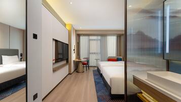 Quarto de hotel com cama, escrivaninha, cadeira, televisão e banheiro com pia e espelho.