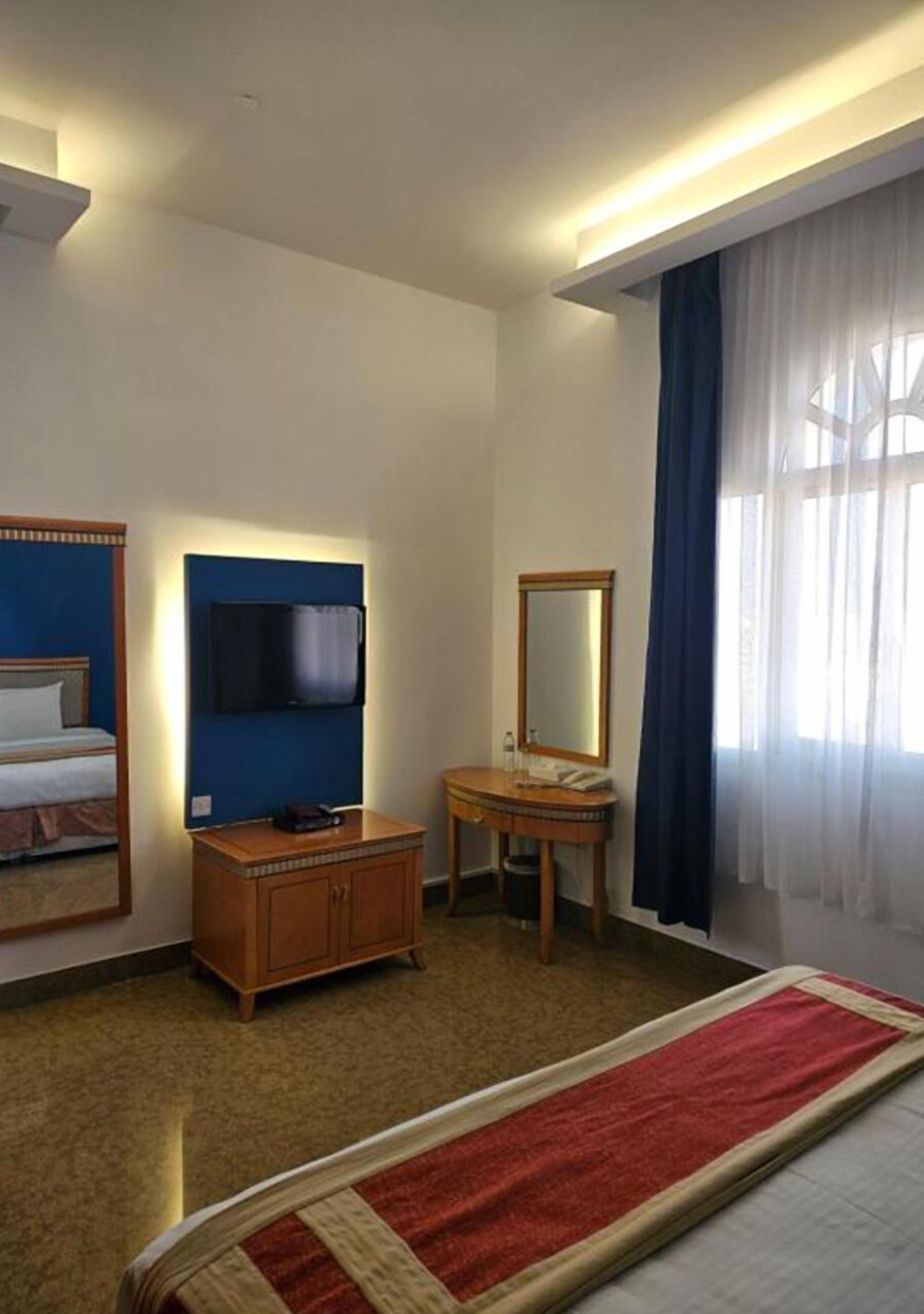 Quarto de hotel com uma cama, televisão na parede, cómoda de madeira, espelho e janela com cortinas.