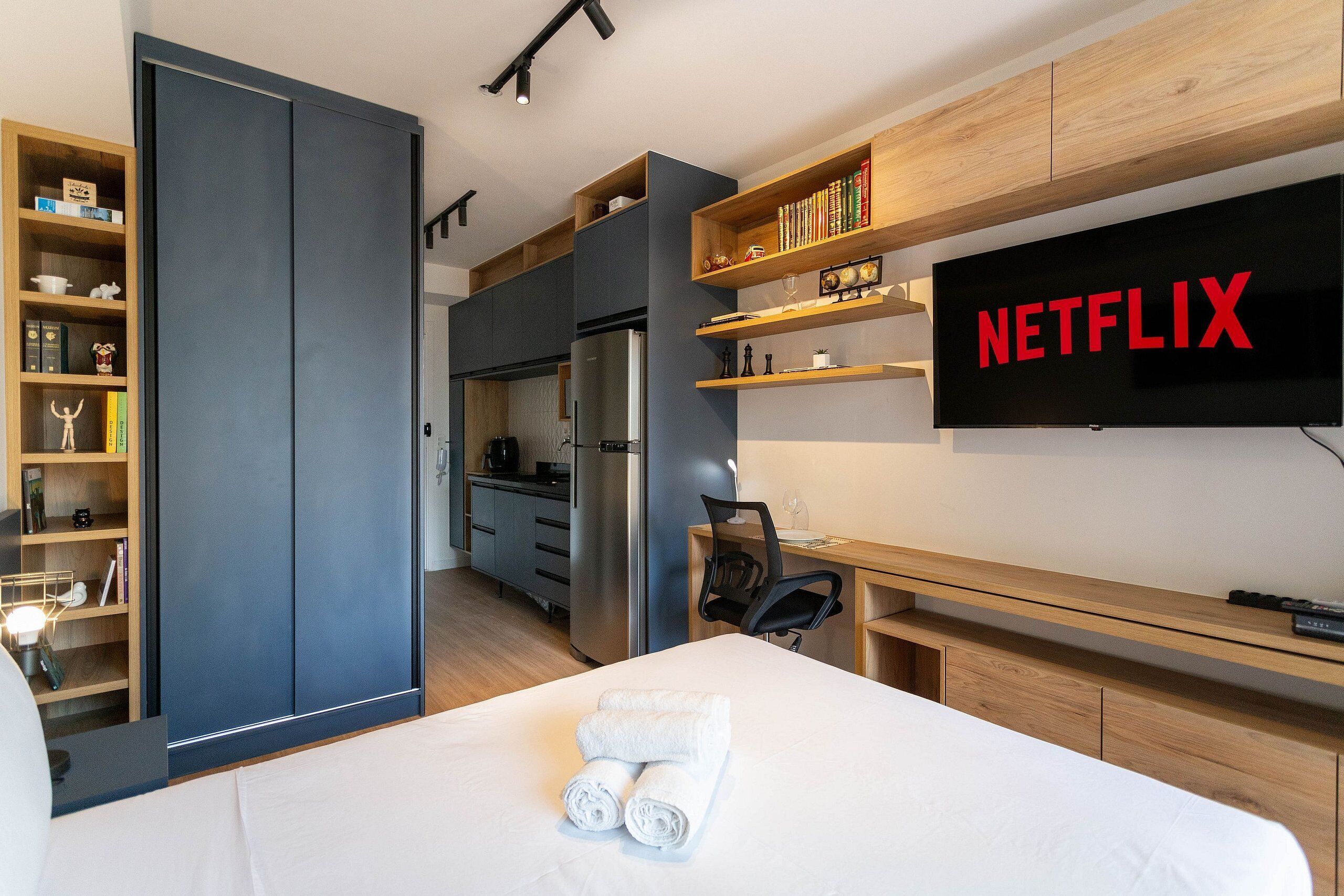 Quarto de hotel moderno com uma cama, uma mesa de escritório, uma televisão e uma kitchenette ao fundo.
