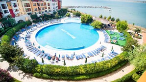 Uma grande piscina circular rodeada por espreguiçadeiras e guarda-sóis, com vista para o mar e um hotel ao fundo.