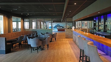 Uma área de bar com balcão de madeira e banquetas, uma sala de estar com sofás e cadeiras, e um mural na parede.