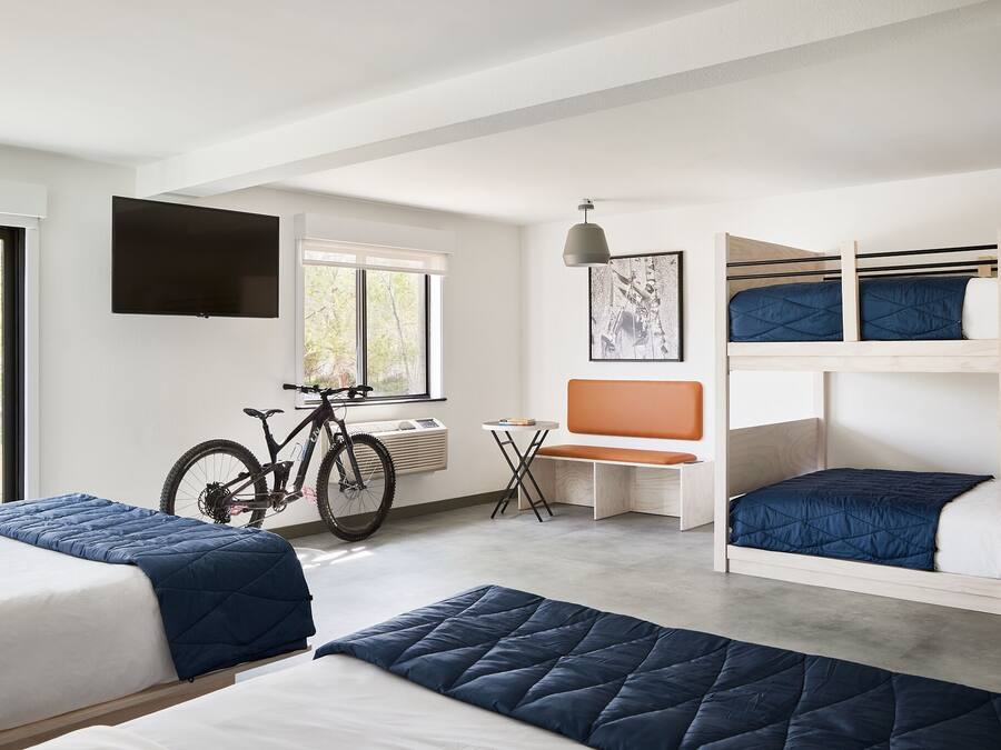Quarto com beliches, uma bicicleta, televisão e janela com vista para árvores.