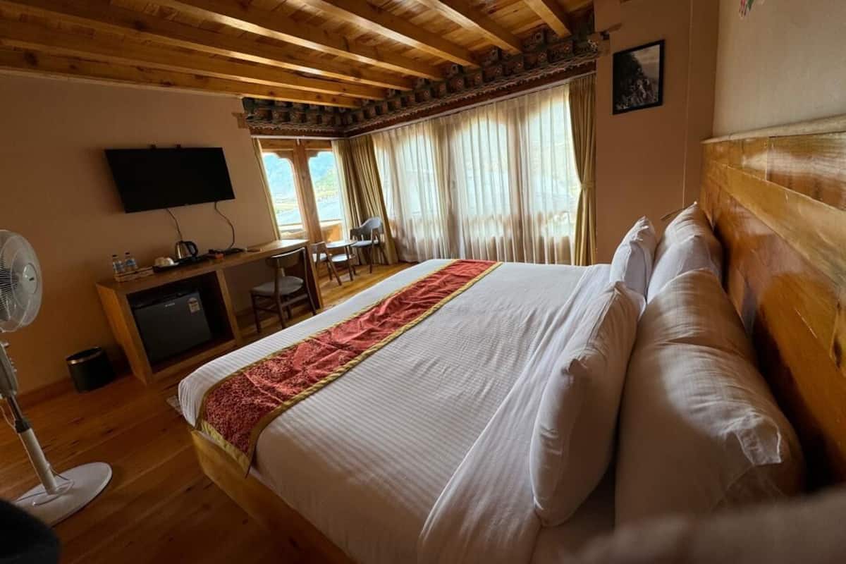 Quarto de hotel com teto de madeira, cama com roupa de cama branca, escrivaninha com computador e televisão na parede.