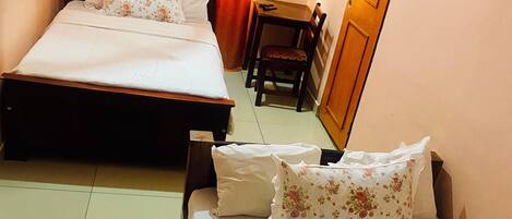Quarto de hotel com duas camas, cada uma com almofadas estampadas com padrão floral, cabeceira de madeira e uma pequena mesa com uma cadeira.