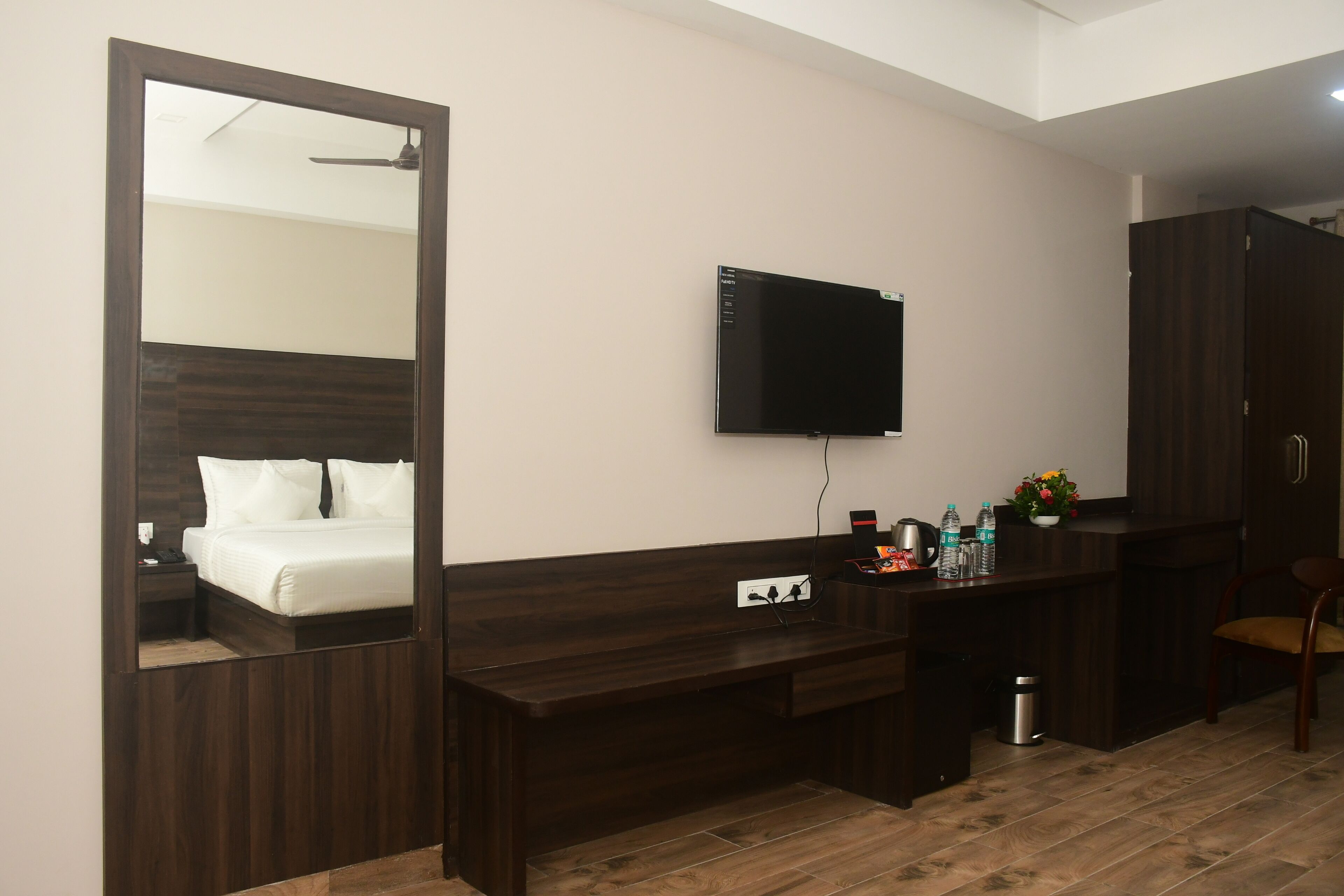 Quarto de hotel com escrivaninha de madeira, televisão de tela plana na parede, espelho e cama com roupa de cama branca.