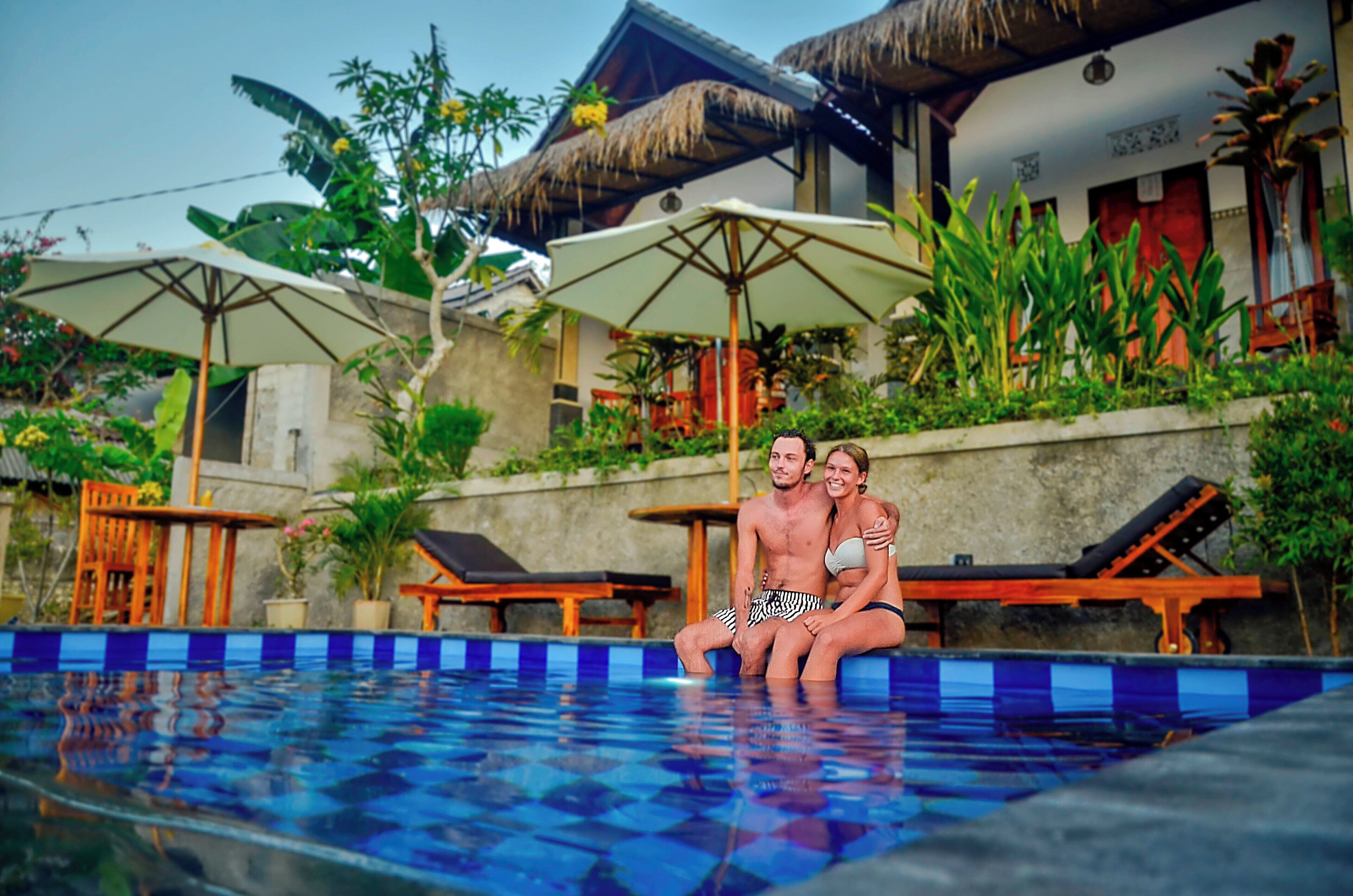 Um casal sentado à beira da piscina, com edifícios de telhado de palha e plantas tropicais ao fundo.