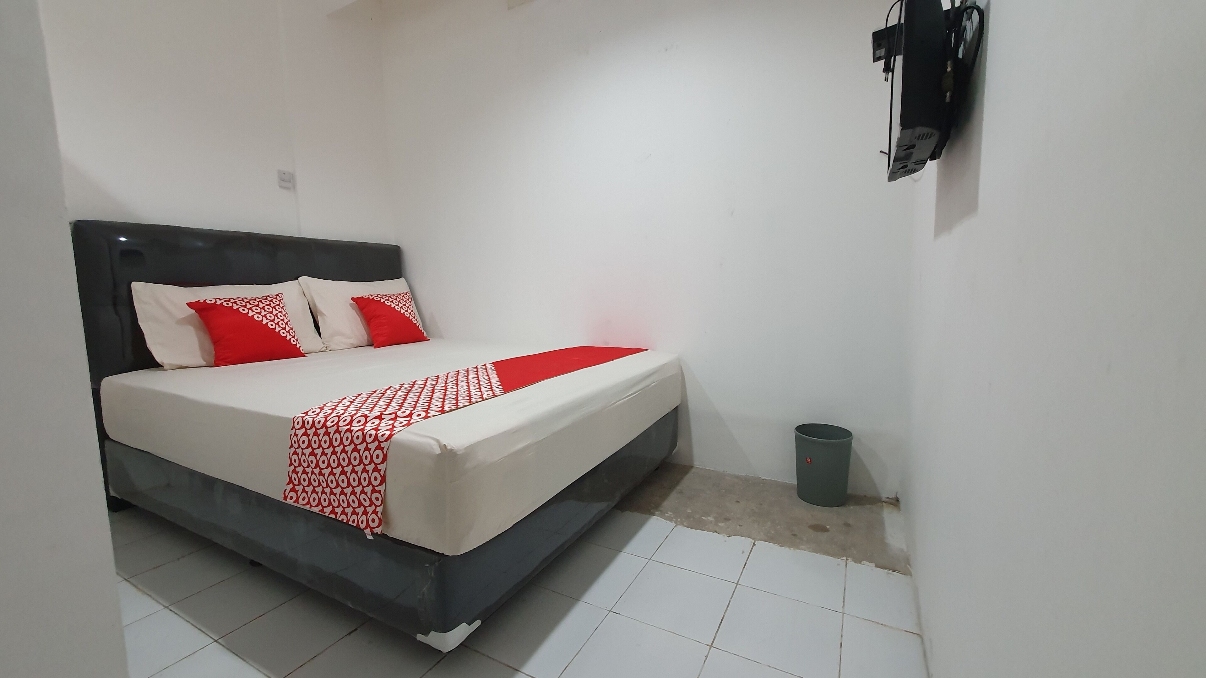 Quarto pequeno e limpo, com uma cama, um cesto de lixo e uma televisão presa na parede.