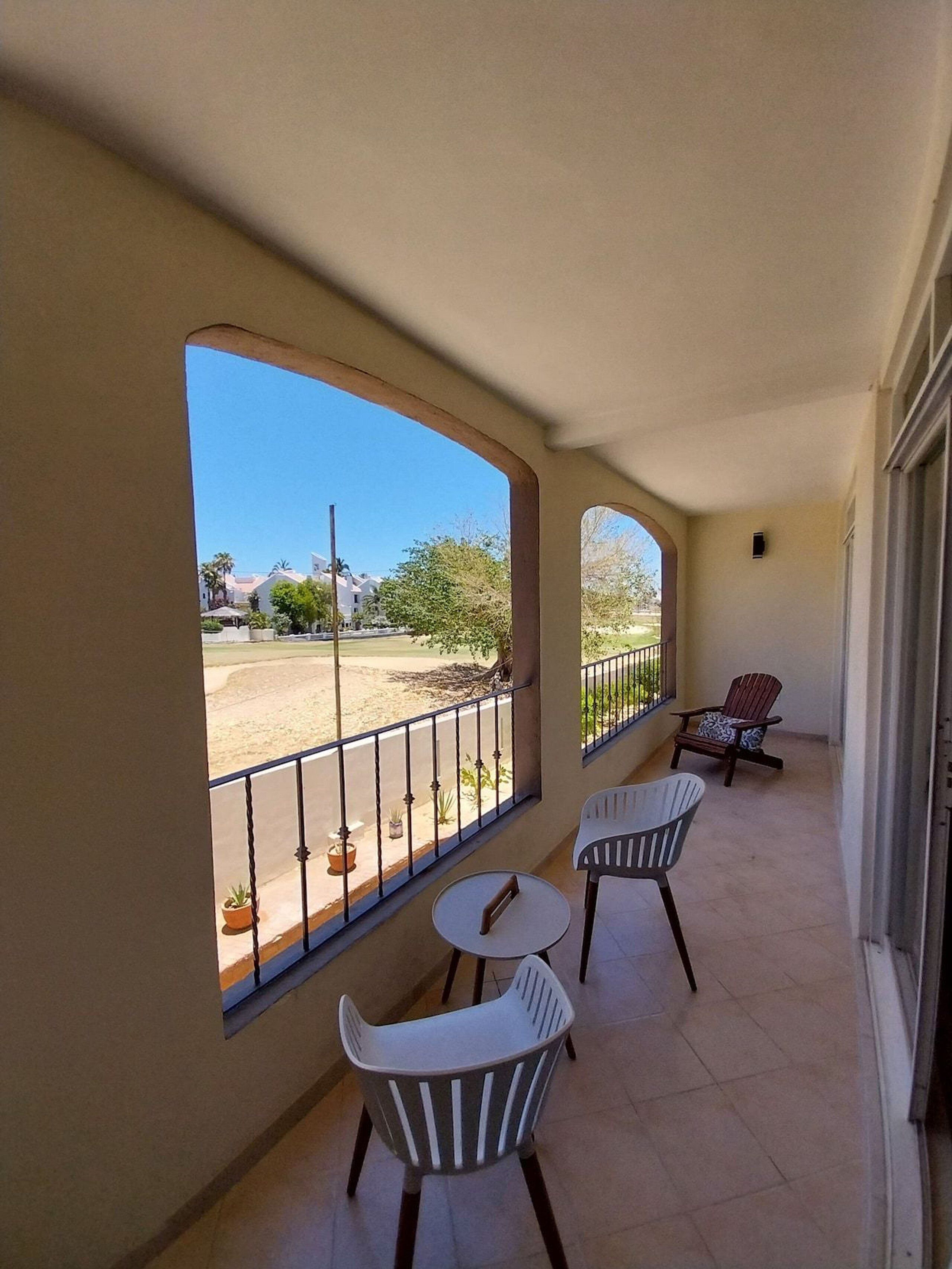 Uma varanda com duas cadeiras e uma mesa pequena, com vista para uma área arenosa e edifícios ao longe.